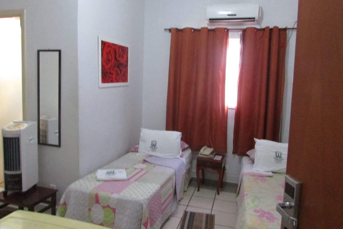 Quarto pequeno e limpo com duas camas de solteiro, uma mesa de cabeceira com abajur, um espelho e uma cortina vermelha cobrindo uma janela.