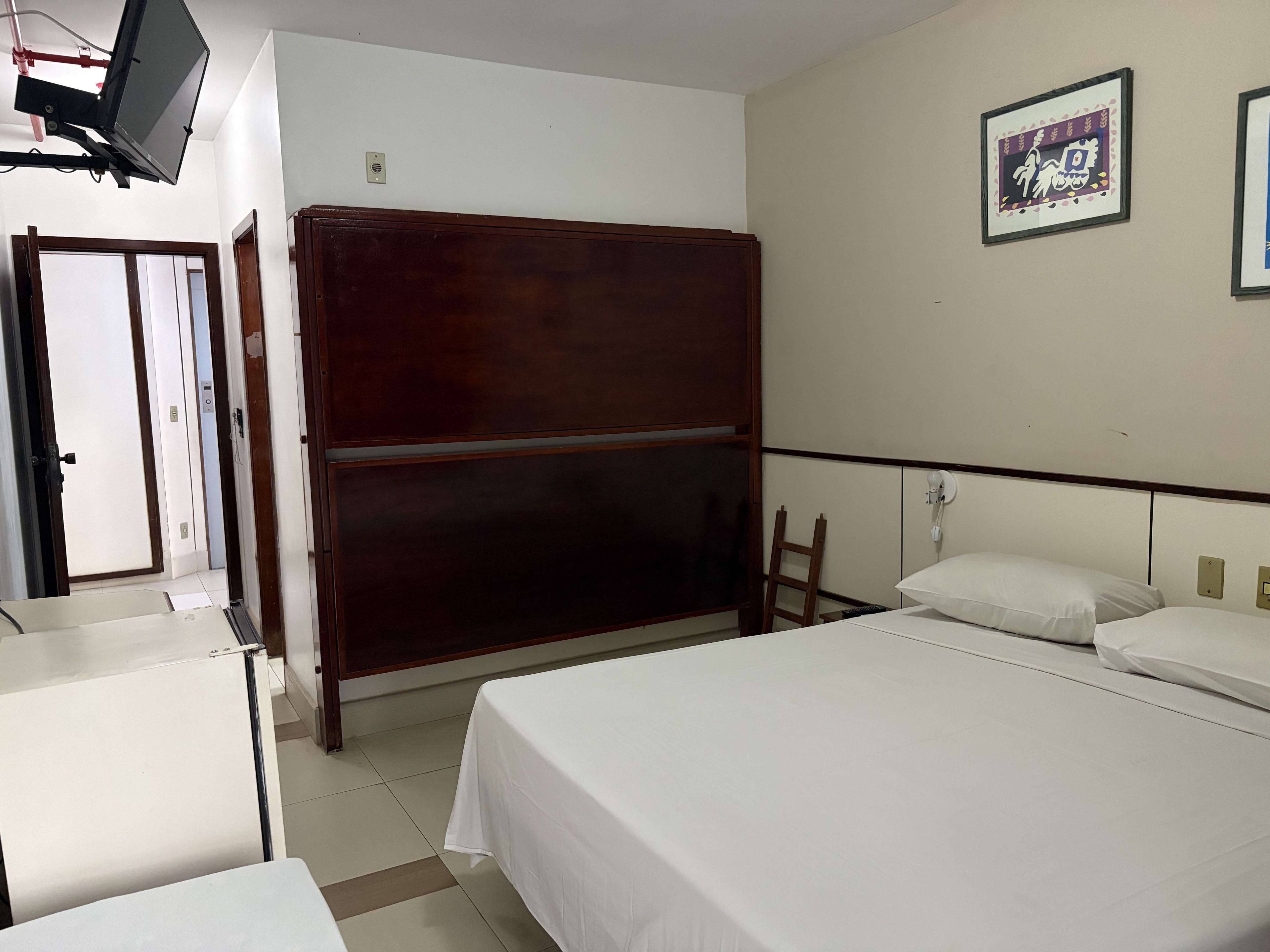 Quarto de hotel com cama, escrivaninha, televisão fixada na parede e porta que dá para outro cômodo.