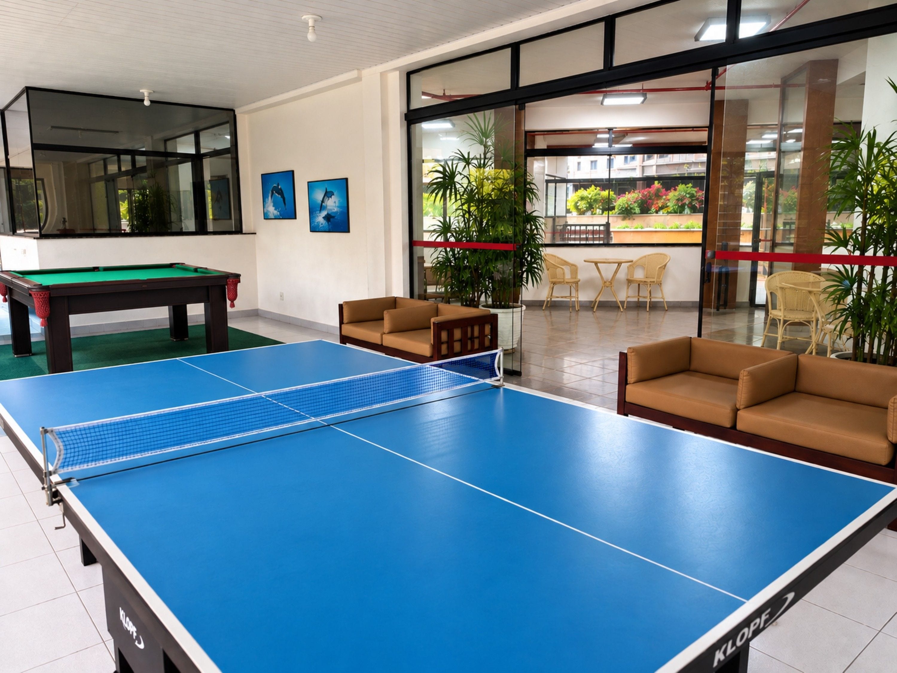 Uma mesa de pingue-pongue em um espaço que também tem uma mesa de bilhar, dois sofás e um painel de vidro com vista para o exterior.