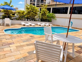 Piscina com espreguiçadeiras e uma mesa, rodeada por piso cerâmico e vegetação.