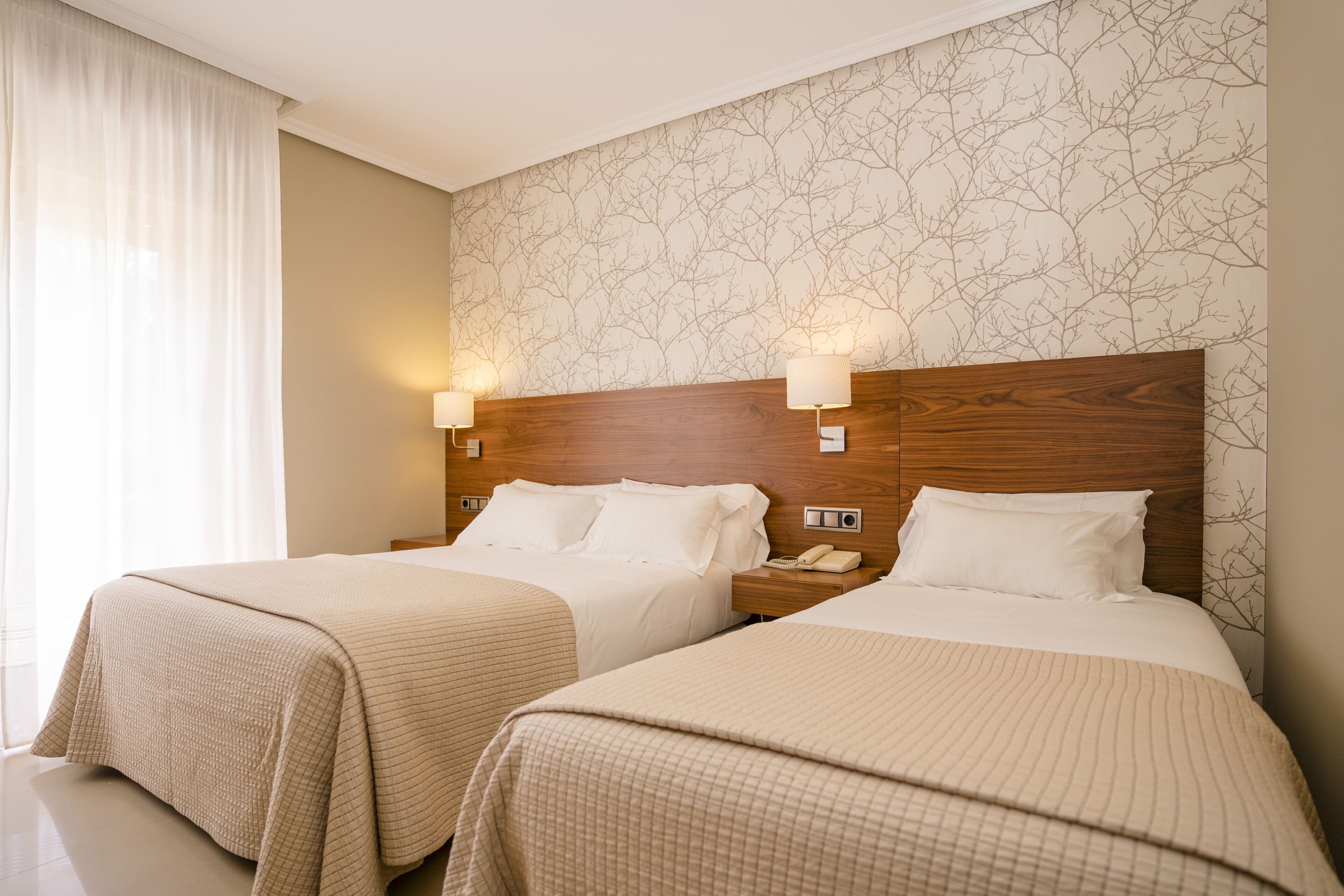Quarto de hotel com duas camas, cabeceira de madeira, abajures e papel de parede estampado.