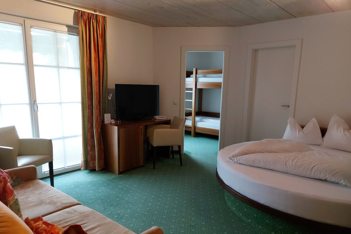 Quarto de hotel com uma cama, uma mesa com televisão, uma cadeira, um sofá e uma beliche ao fundo.