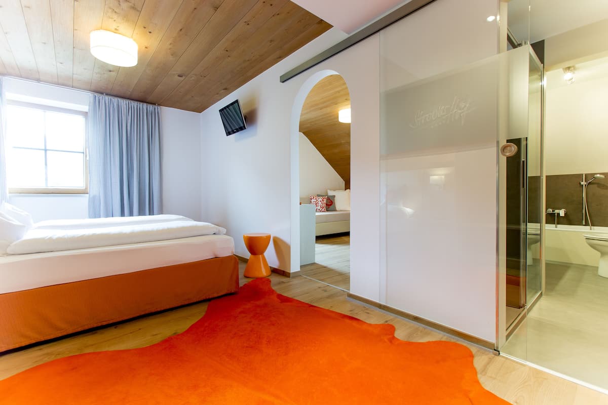 Quarto moderno com cama, tapete laranja grande e banheiro visível através de uma porta de vidro.
