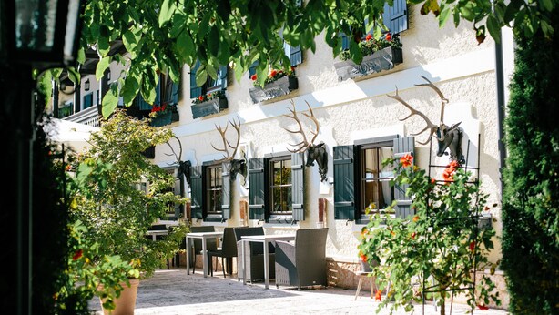 Uma área de estar externa com mesas e cadeiras, um edifício branco com persianas verdes e chifres decorativos na parede.