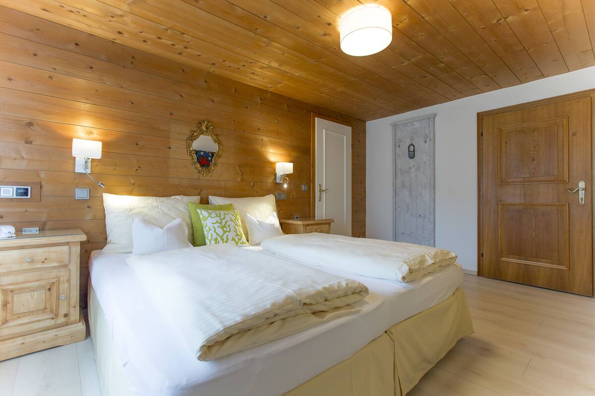 Uma cama de casal com lençóis brancos, duas mesas de cabeceira, um criado-mudo e um teto de madeira, além de uma porta.