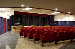 Um teatro com fileiras de assentos vermelhos, um palco com um piano de cauda e um pano de fundo de cortina vermelha.