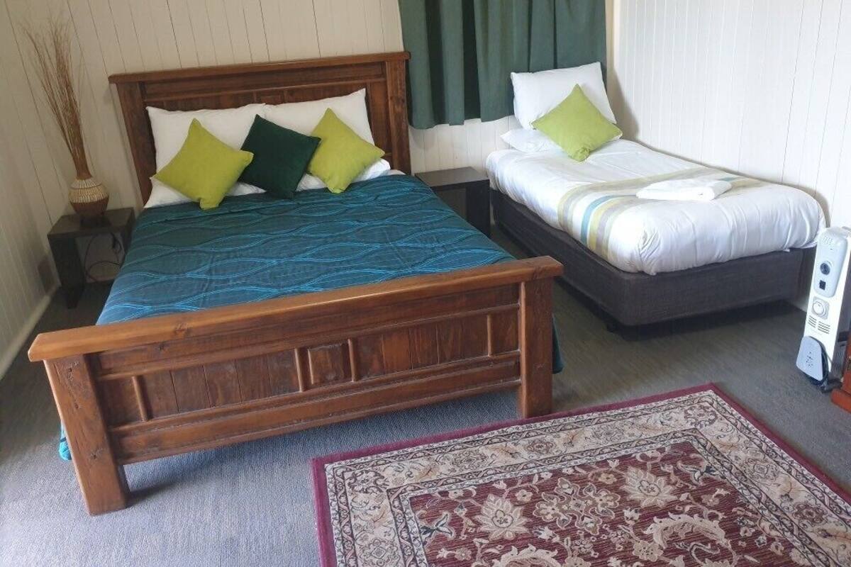 Quarto com duas camas, uma cama de madeira maior e outra menor, ambas com almofadas e mesa de cabeceira.