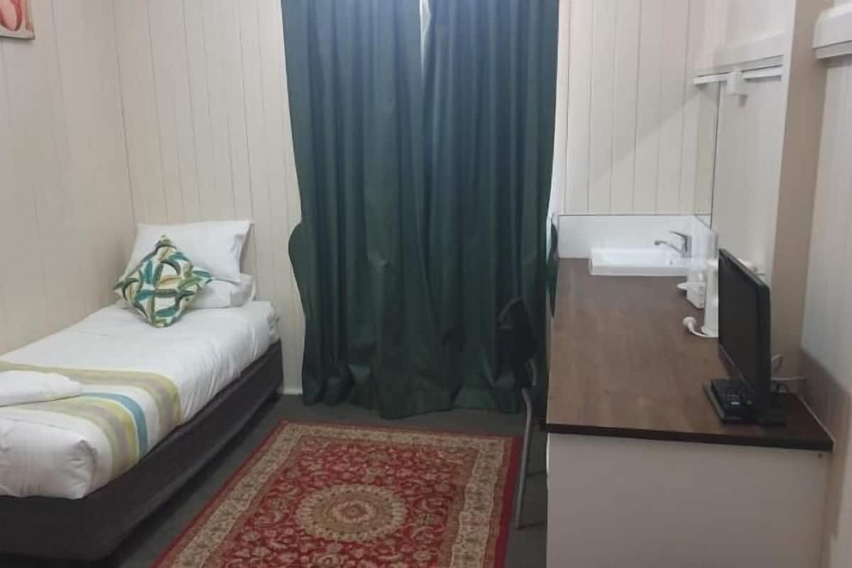 Quarto pequeno com cama, escrivaninha, cadeira, televisão e banheiro.