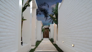 Um caminho estreito ladeado por paredes brancas que leva a uma porta de madeira, com pequenas luzes ao longo do percurso e plantas em ambos os lados.