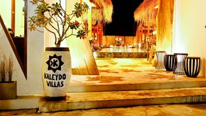 Vista noturna de um resort com entrada de telhado de palha, um grande vaso decorativo com o texto 'Kaleido Villas' e várias mesas com padrão preto e branco.