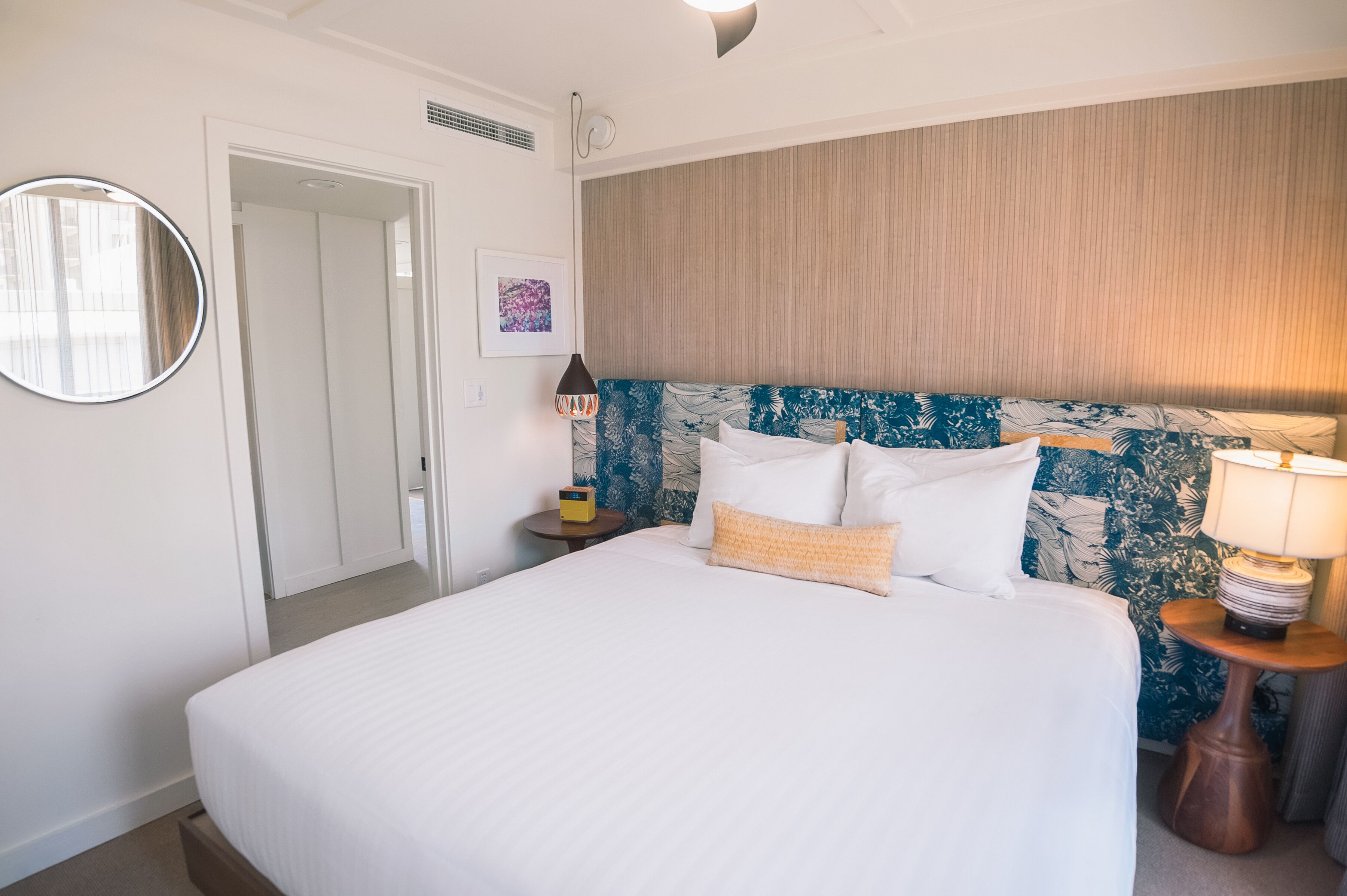 Uma cama bem arrumada com lençóis brancos, cabeceira de madeira com padrão floral azul, um espelho redondo na parede e uma pequena mesa de cabeceira com uma luminária.