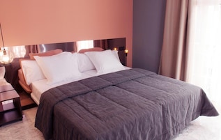 Uma cama bem arrumada com um cobre-leito cinza escuro e almofadas brancas, uma mesa de cabeceira com um abajur, e uma janela com cortinas.