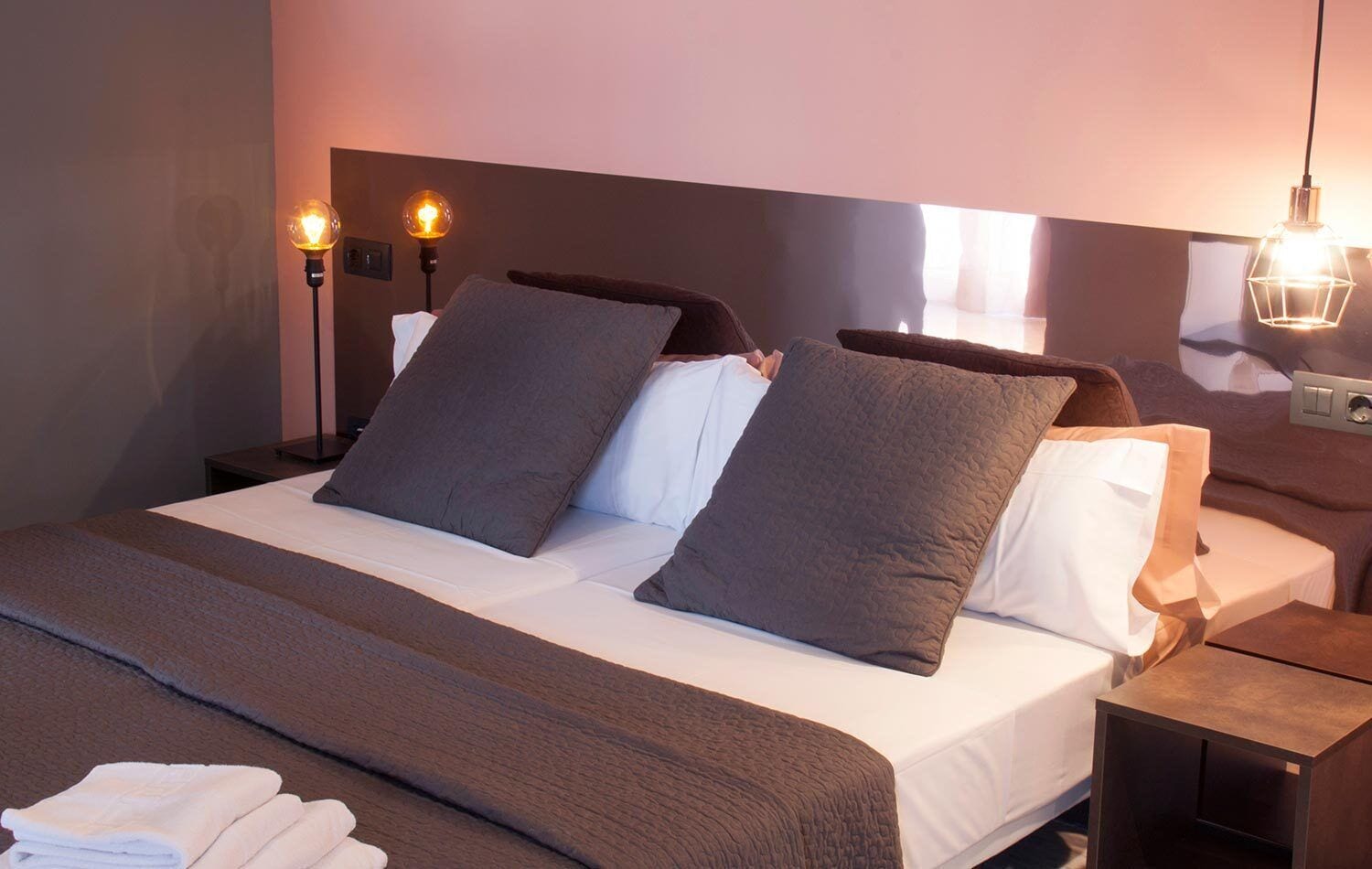 Uma cama bem arrumada com cabeceira escura, duas mesas de cabeceira com abajures e uma luminária na parede.