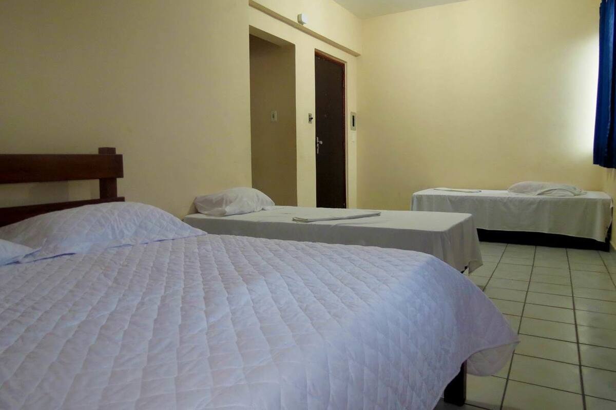 Quarto de hotel com duas camas de solteiro, cabeceira de madeira, uma porta e piso de azulejos.