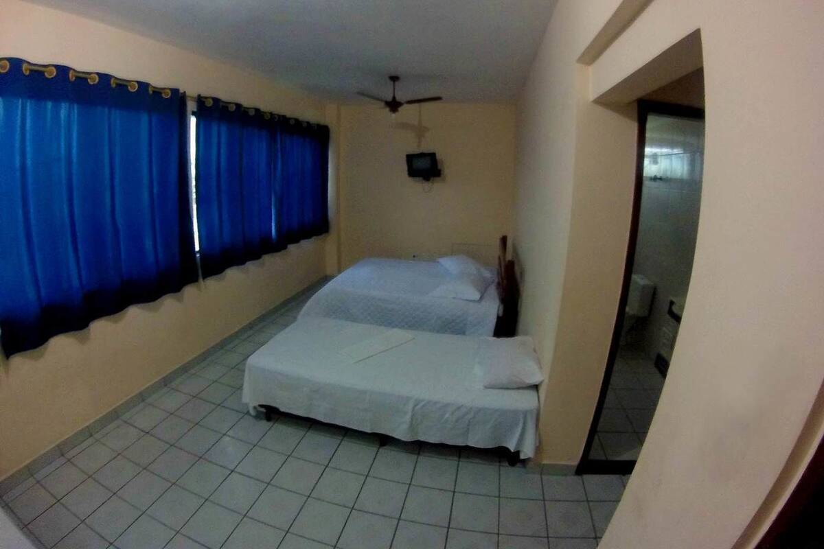 Quarto pequeno e simples com cama, ventilador de teto e cortinas azuis.