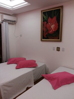 Quarto de hotel com duas camas, cada uma com almofada rosa, um quadro de uma flor vermelha na parede e um aparelho de ar condicionado.