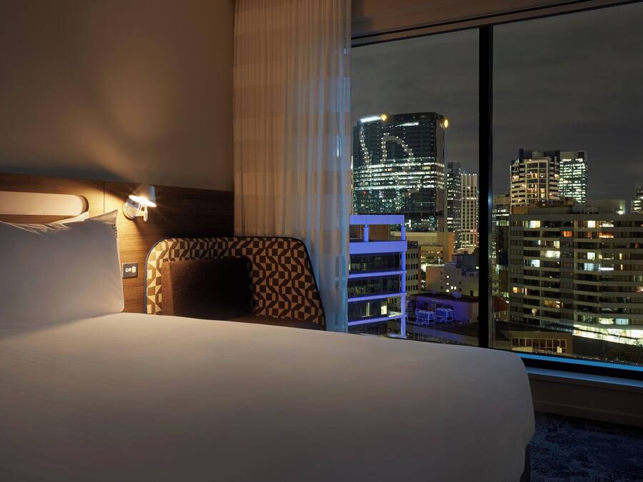 Quarto de hotel com uma cama grande, uma mesa de cabeceira e vista para a paisagem urbana à noite.
