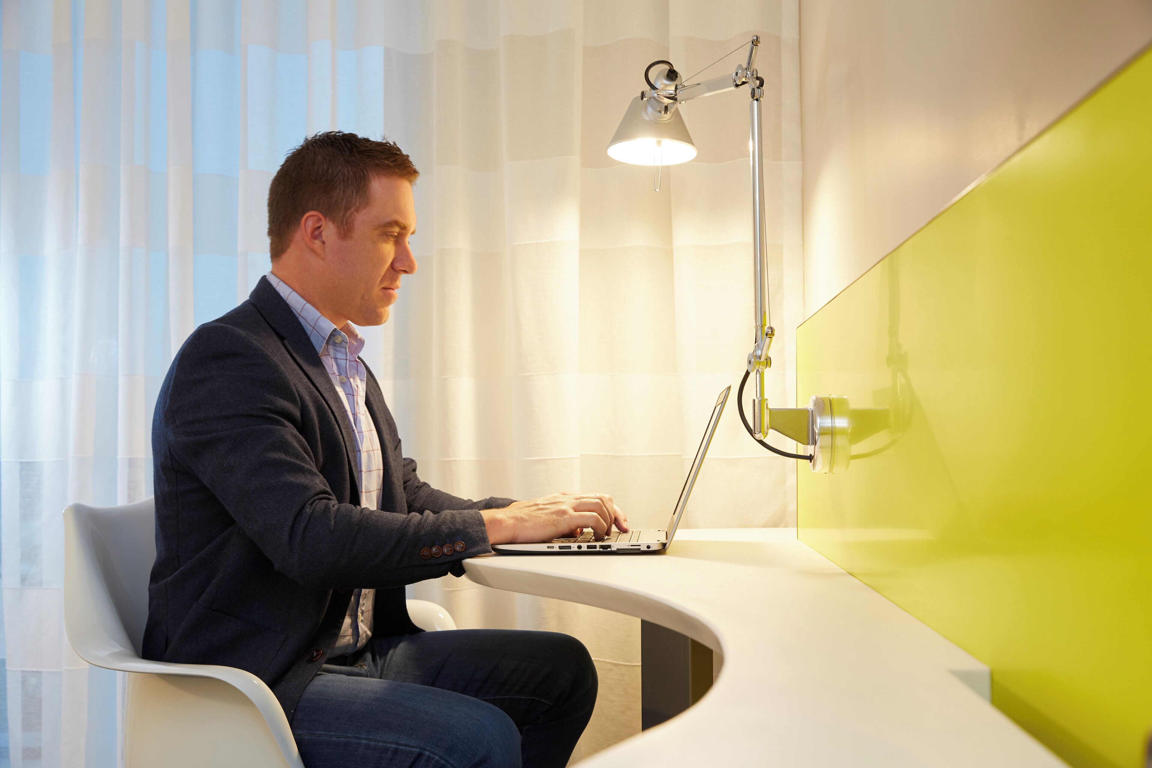 Um homem sentado à mesa com um laptop, uma luminária de mesa e uma escrivaninha branca em um quarto com parede branca e amarela.