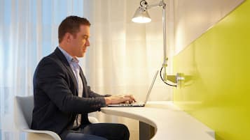 Um homem sentado à mesa com um laptop, uma luminária de mesa e uma escrivaninha branca em um quarto com parede branca e amarela.