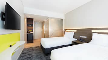 Quarto de hotel com duas camas, uma escrivaninha, televisão de tela plana, minibar e banheiro com chuveiro e vaso sanitário.
