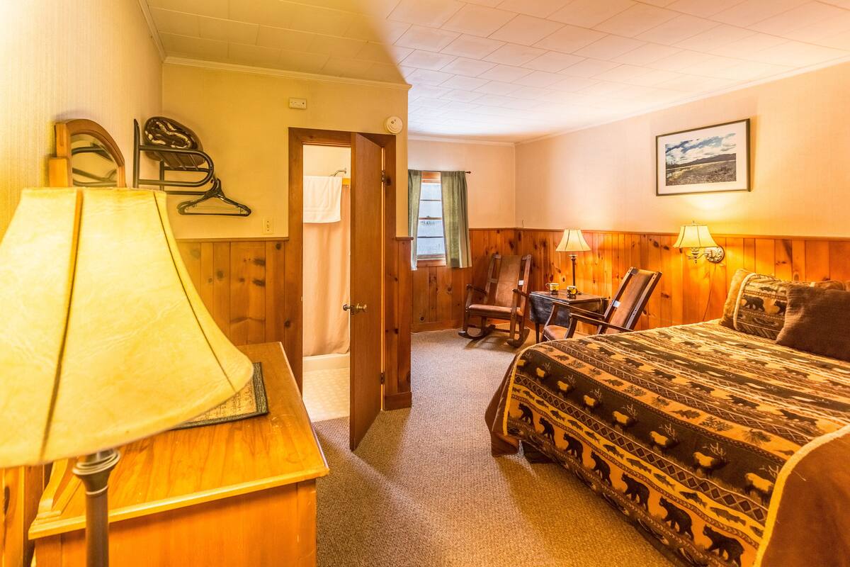 Quarto de hotel com cama, escrivaninha com luminária, cadeira, porta de madeira e janela com cortinas.