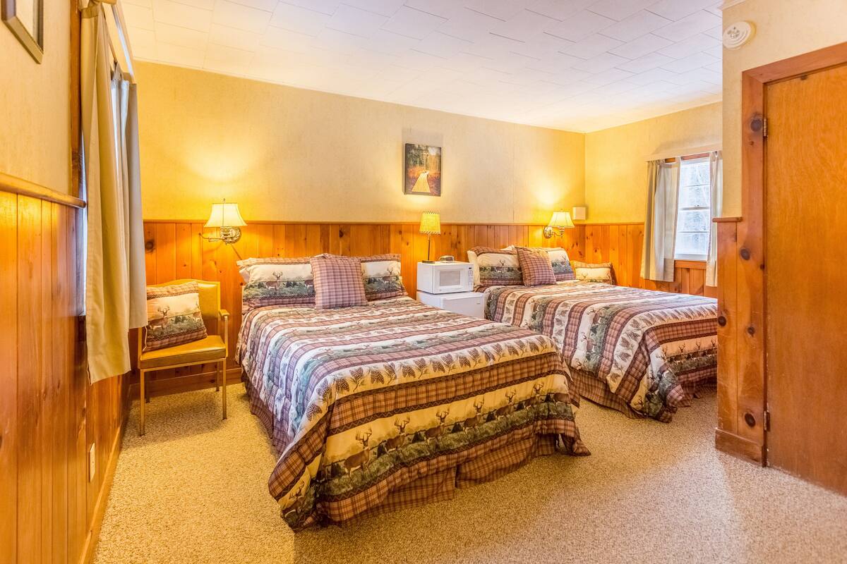 Quarto com duas camas, paredes de madeira, teto com padrão quadriculado e uma única janela com cortinas.