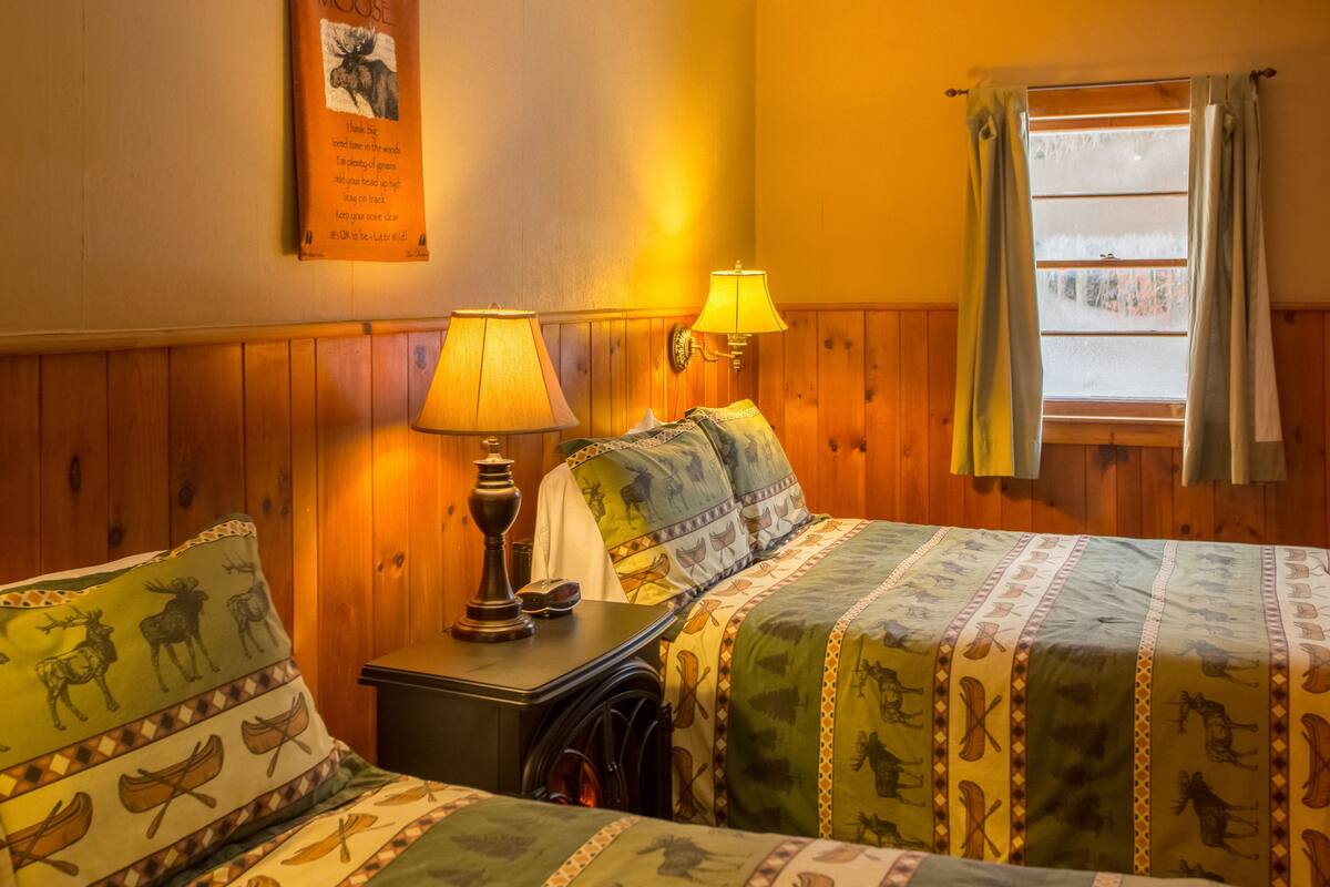 Quarto com duas camas, paredes de madeira, janela com cortinas e luminária fixada na parede.