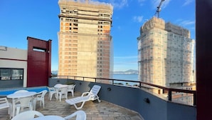 Terraço no telhado com mobiliário de plástico branco, piscina e vista para um horizonte urbano com edifícios em construção.