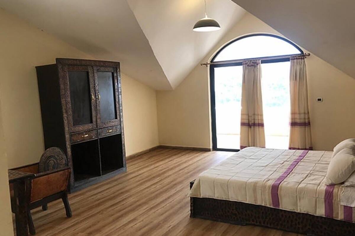 Quarto com piso de madeira, cama com colcha estampada, guarda-roupa e uma janela grande com cortinas.