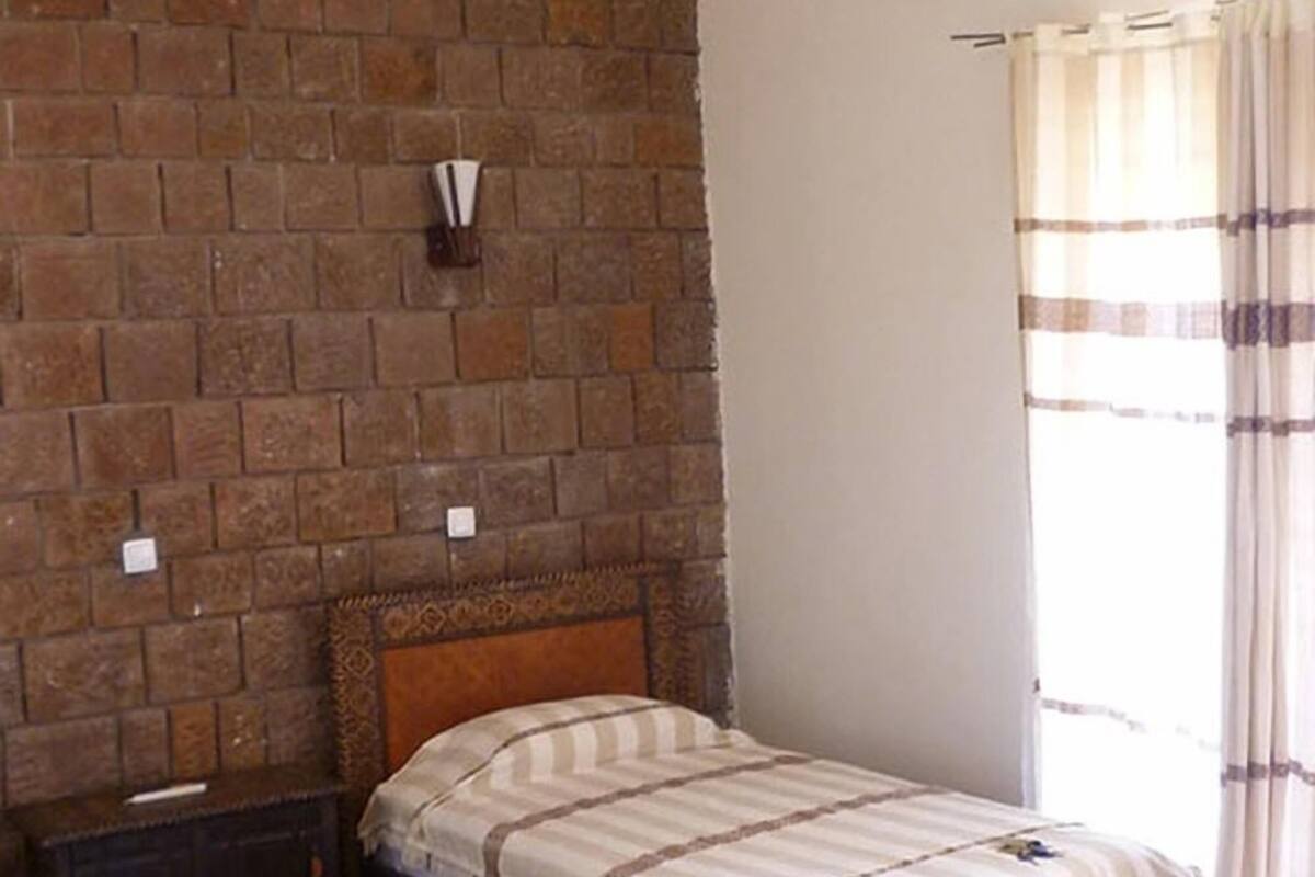 Quarto com parede de tijolos, cama com colcha listrada, mesa de cabeceira e janela com cortinas.