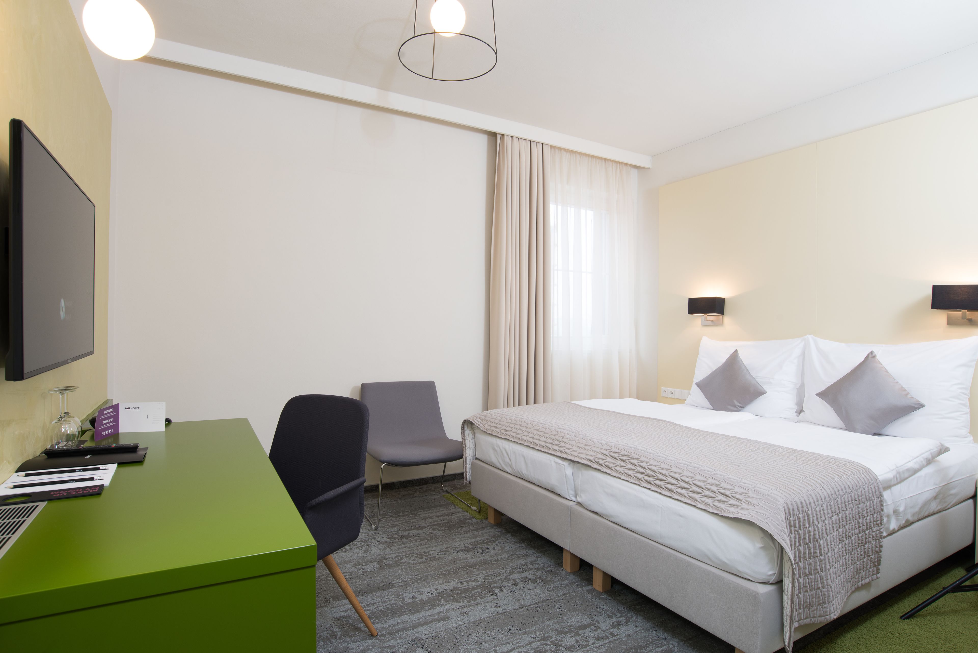Quarto de hotel com uma cama, escrivaninha, cadeira, televisão e janela com cortinas.