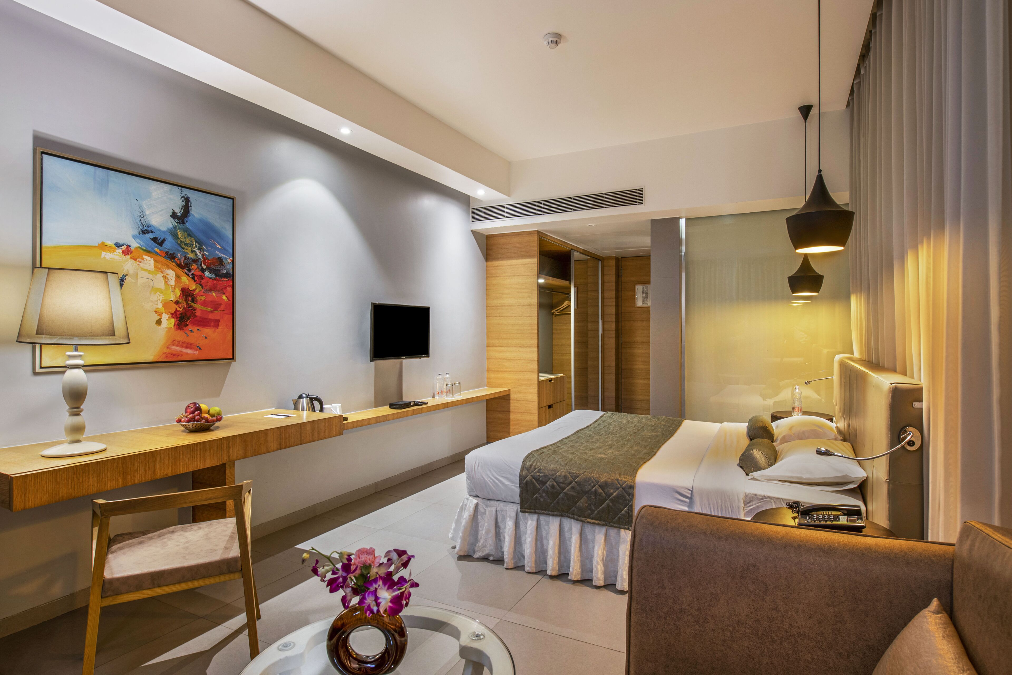 Quarto de hotel moderno com uma cama, uma escrivaninha com luminária, uma televisão e um quadro grande na parede.
