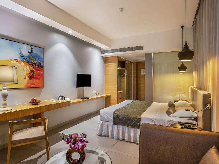 Quarto de hotel moderno com uma cama, uma escrivaninha com luminária, uma televisão e um quadro grande na parede.