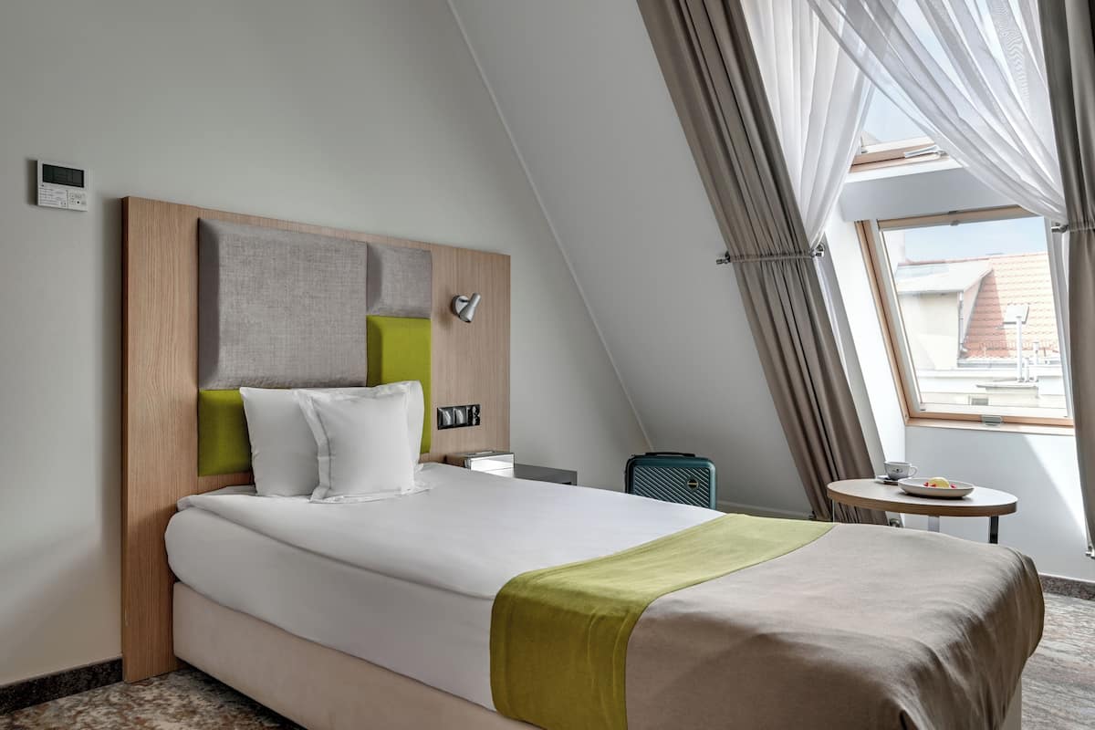 Quarto de hotel com cama, mesa de cabeceira, pequena mesa com xícara e prato, e janela com cortinas.