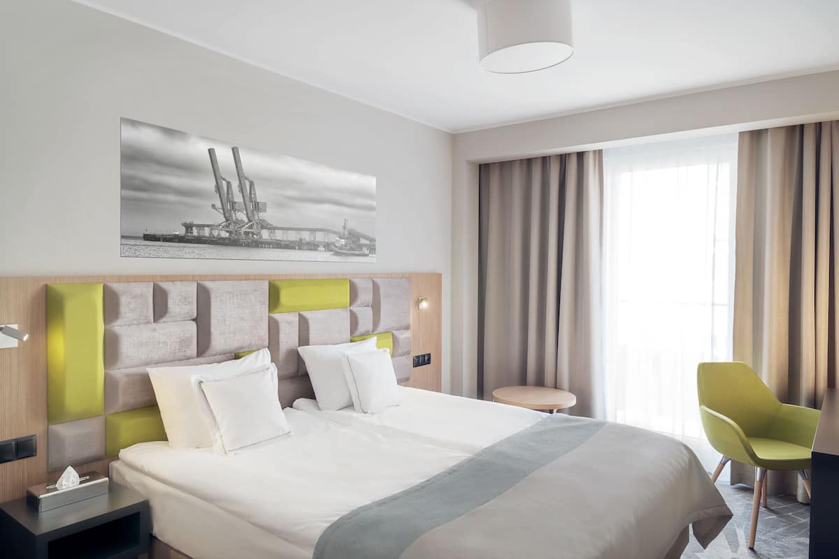 Quarto de hotel com uma cama grande, cabeceira com padrões geométricos, mesa de cabeceira, uma cadeira, uma mesinha redonda e uma janela com cortinas.