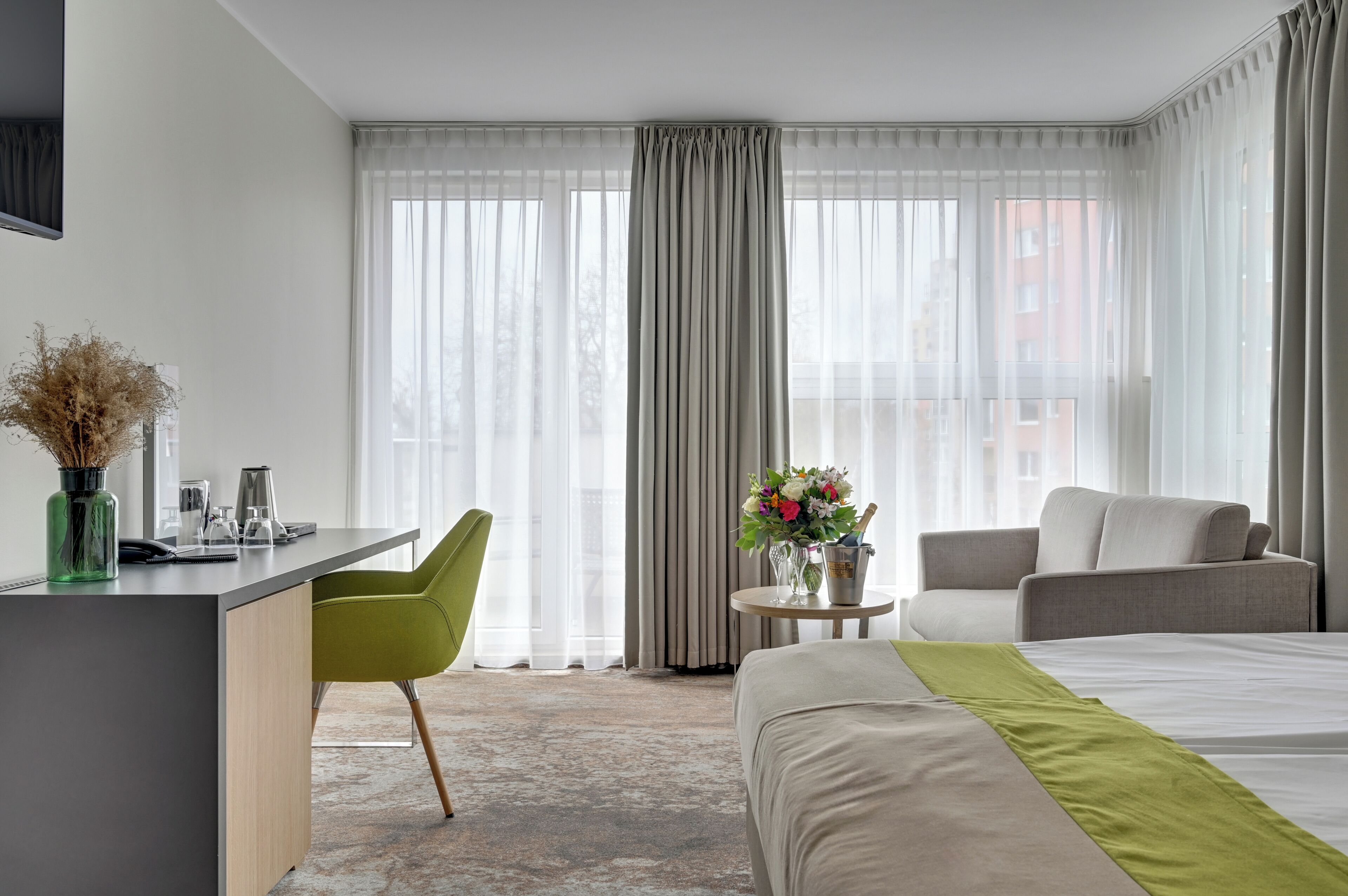 Quarto de hotel com cama, escrivaninha, cadeira, mesinha de centro com um vaso de flores e grandes janelas com cortinas.