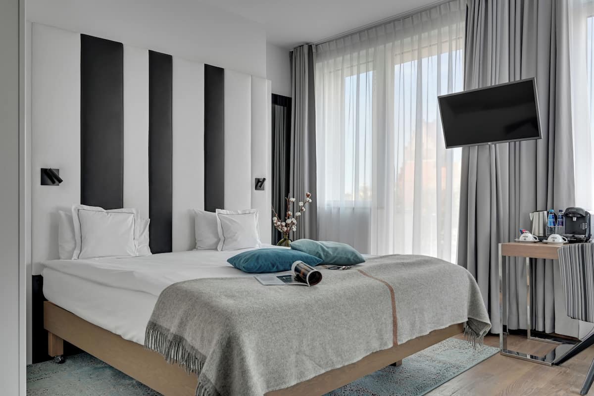 Quarto de hotel moderno com uma cama grande, televisão de tela plana, escrivaninha com cadeira e vista para a cidade através de grandes janelas.