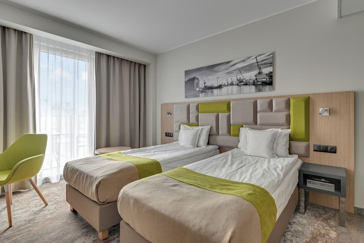 Quarto de hotel com duas camas, uma cadeira verde, um quadro na parede e uma mesinha de cabeceira.