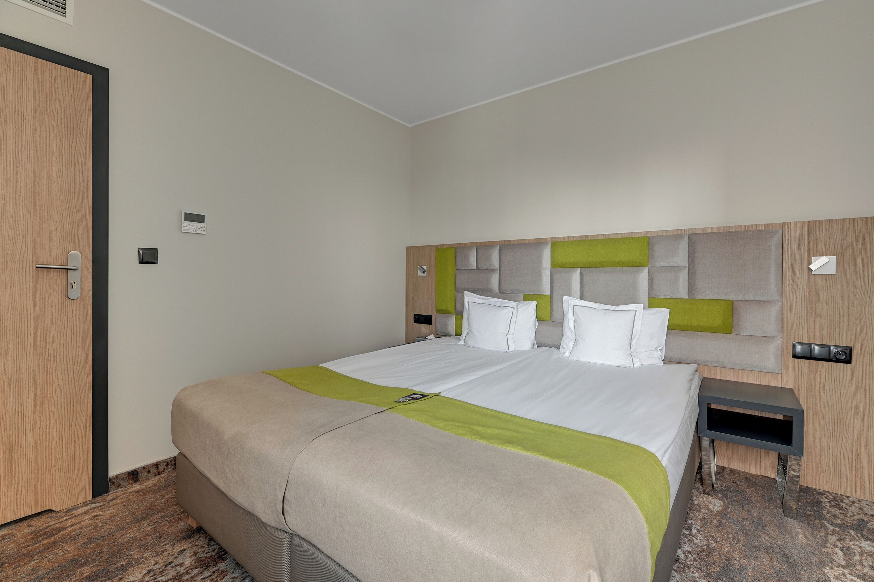 Quarto de hotel com cama, mesa de cabeceira, porta e painel de controle fixado na parede.