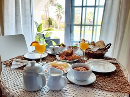 Uma mesa de café da manhã com um laptop, um bule, uma jarra, uma tigela de cereal, croissants, frutas e uma cesta de pão.