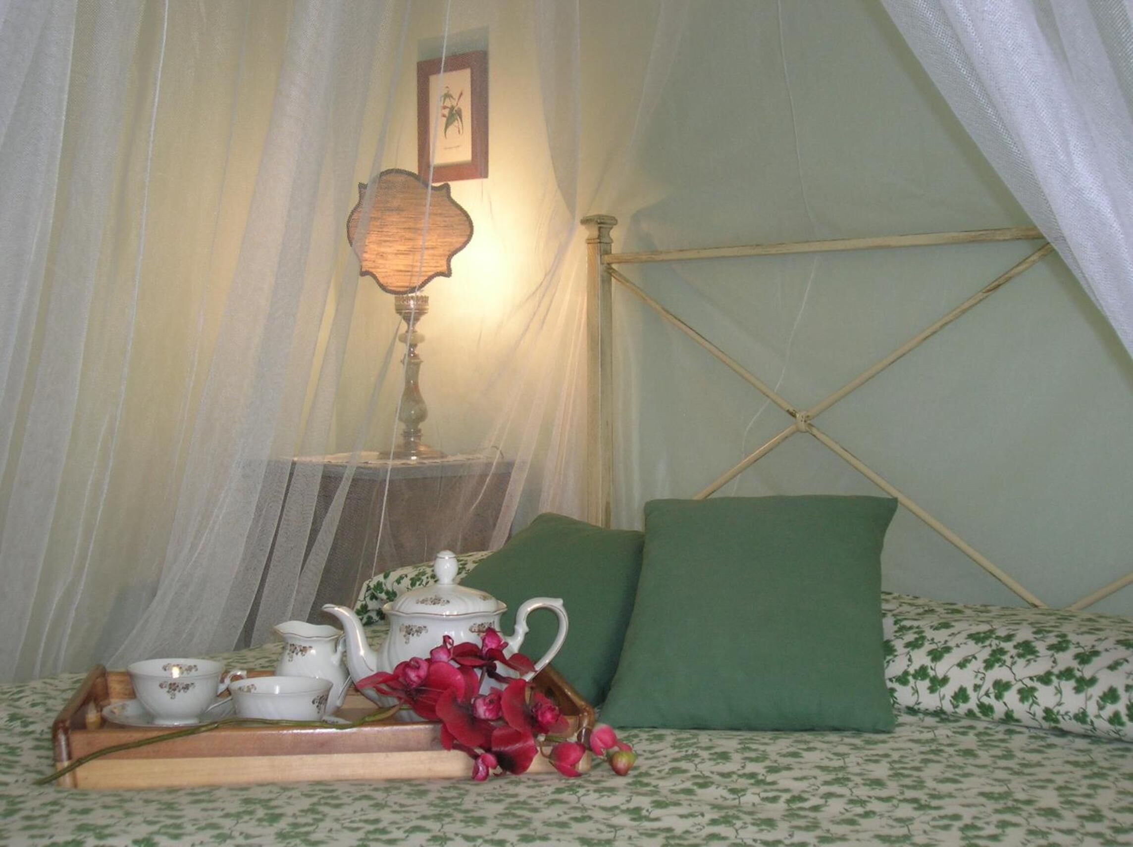 Quarto aconchegante com cama, mesa de cabeceira com abajur e bandeja com chá e flores.