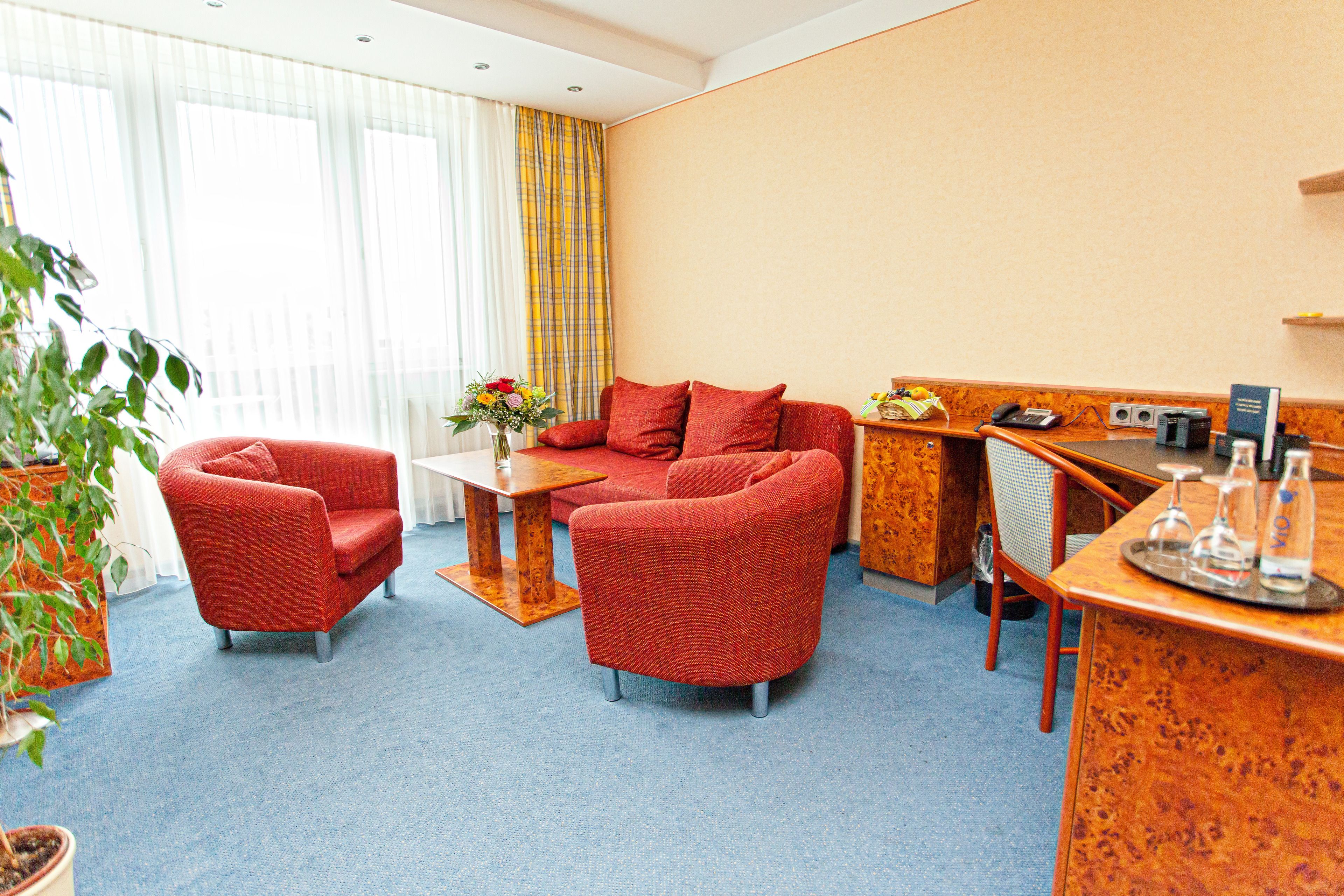 Quarto de hotel com sofá vermelho, pequena mesa de madeira com um vaso de flores, minibar e uma grande janela com cortinas.