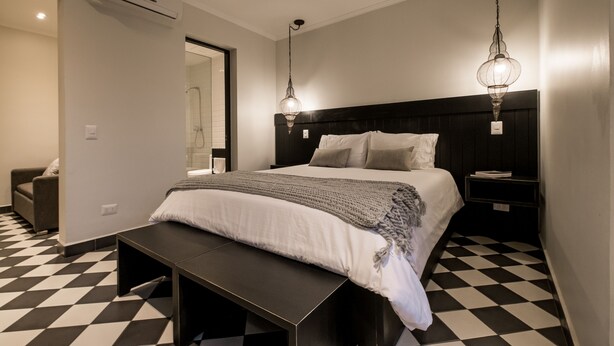 Quarto moderno com piso xadrez preto e branco, uma cama grande com cobertor cinza, duas mesas de cabeceira e uma luminária pendente.