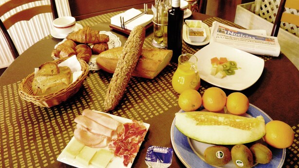 Mesa de café da manhã com croissants, pão, queijo, presunto, frutas e suco.