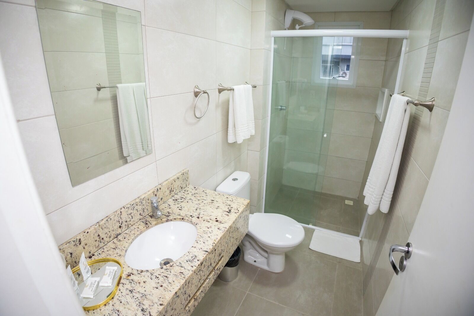 Casa de banho com bancada de granito, duas pias, sanita e cabine de duche com parede de vidro.