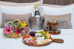 Uma mesa com um arranjo de flores, uma garrafa de champanhe, uma tábua de queijos e uma bandeja de morangos cobertos de chocolate.