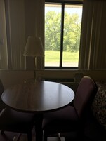 Quarto com uma mesa redonda, uma cadeira, um abajur e uma janela com vista para árvores.