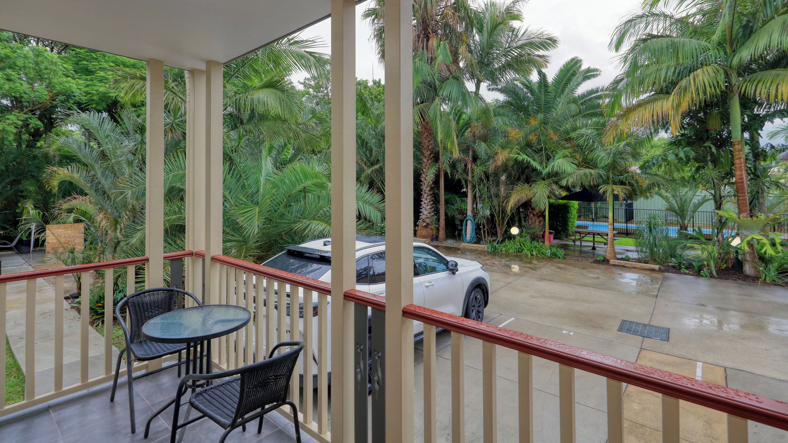 Varanda com mesa e cadeiras, com vista para um estacionamento, um carro branco e vegetação exuberante.