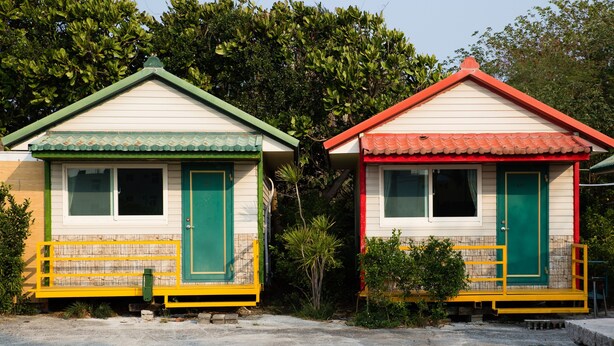 Duas cabanas pequenas e coloridas, cada uma com portas e janelas verdes, além de um pequeno alpendre, em meio à vegetação.
