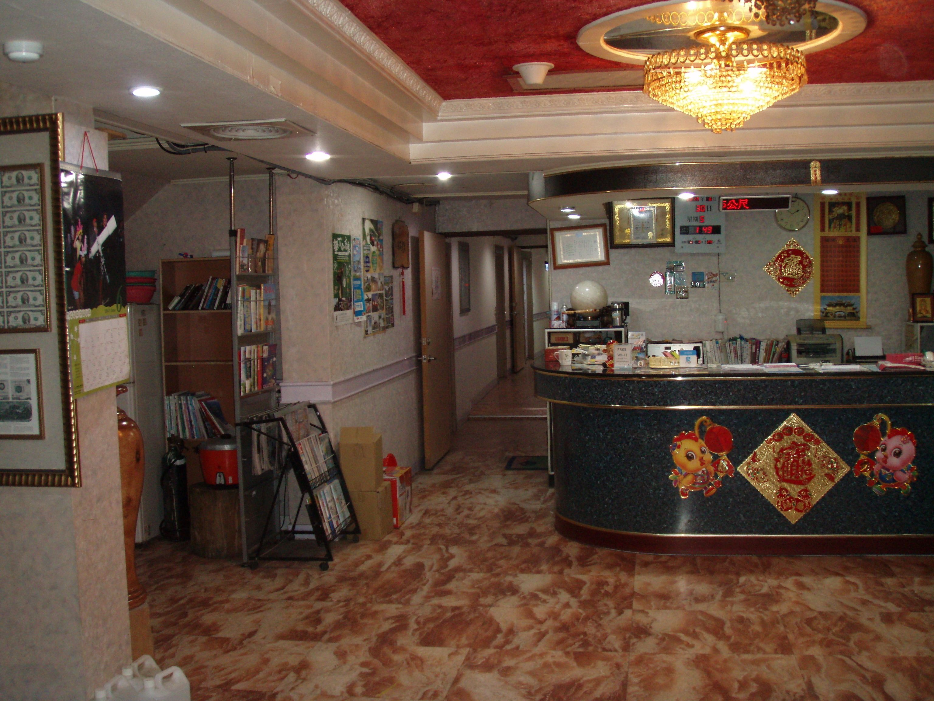 Uma área de recepção de hotel com piso de mármore, balcão de atendimento decorado, um lustre e diversas decorações nas paredes, incluindo calendários e quadros.